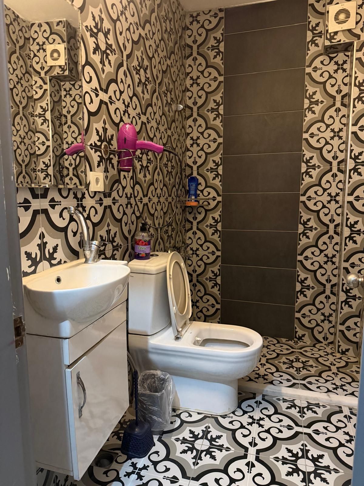 Casa de banho com papel de parede estampado, uma pia branca, sanita e zona de duche.