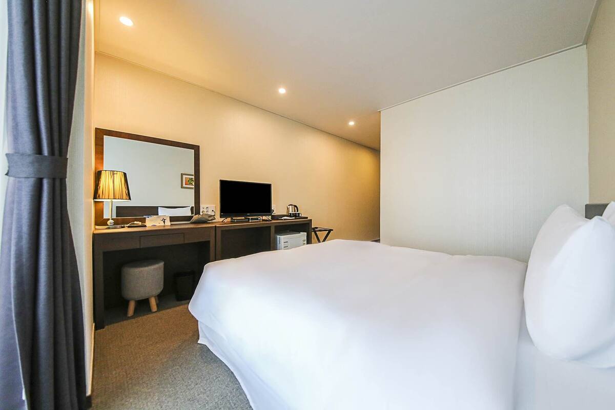 Quarto de hotel com cama, escrivaninha, televisão e espelho.