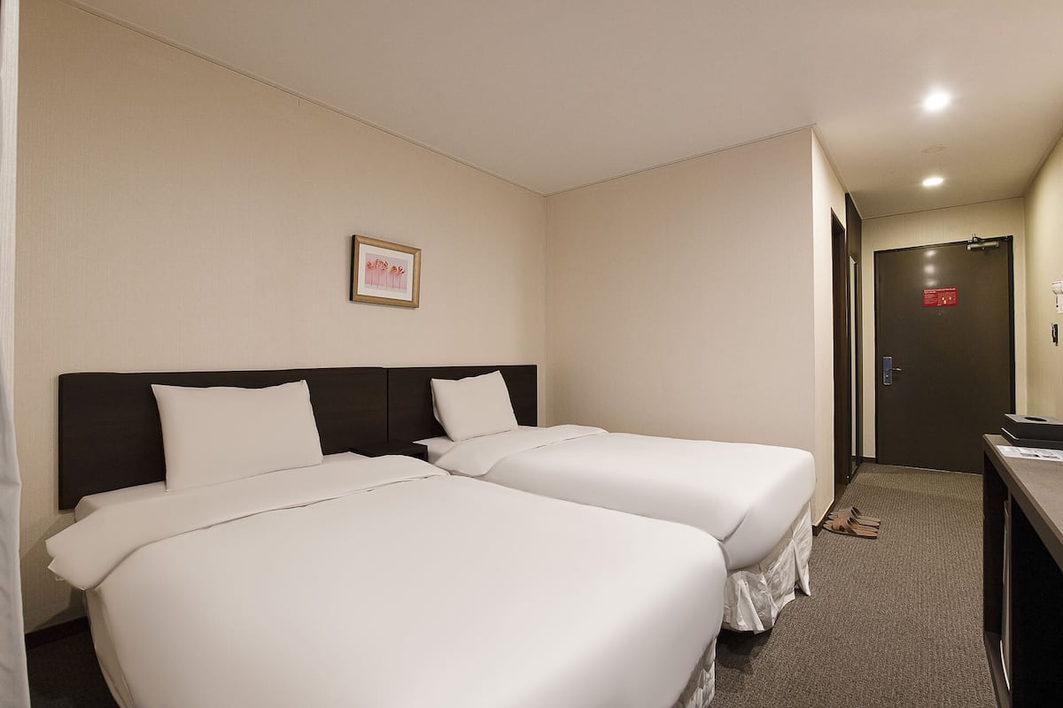 Quarto de hotel com duas camas, um quadro na parede, uma porta com uma placa vermelha e uma mesa de escritório com um computador.