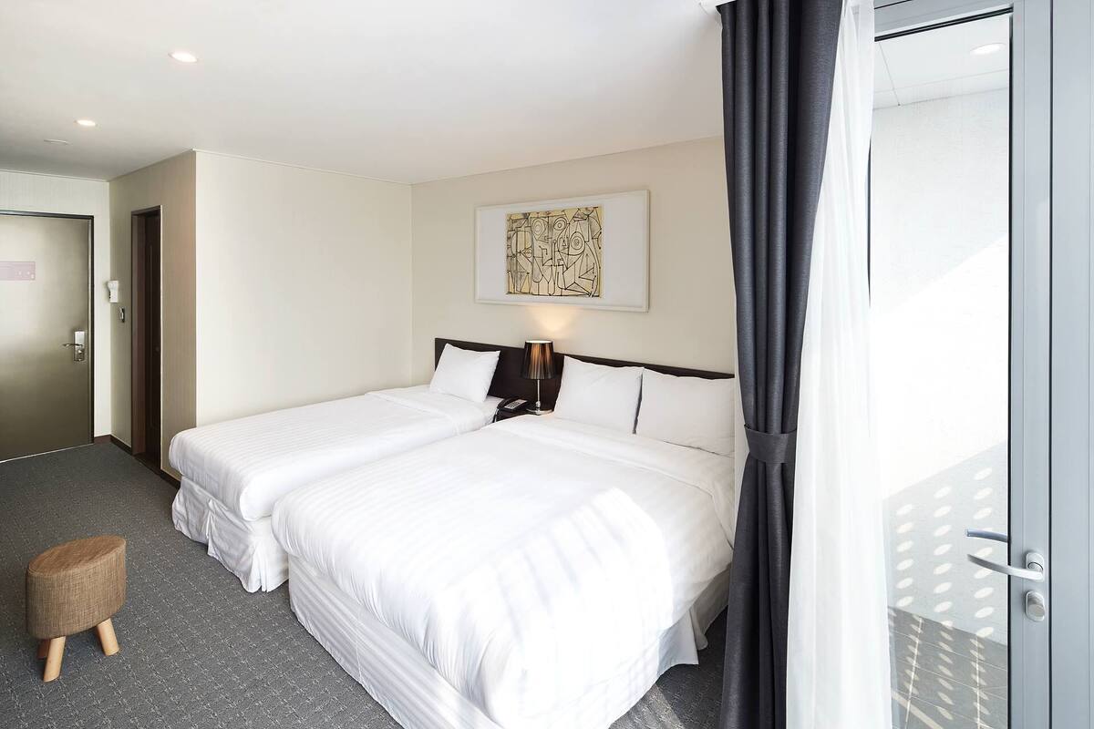 Quarto de hotel com duas camas, um banquinho pequeno, um quadro na parede e uma porta que dá para outro aposento.