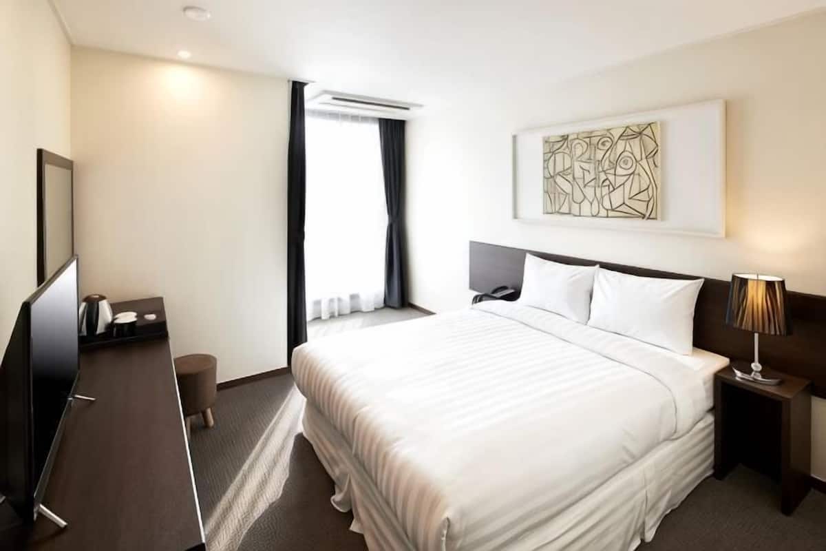 Quarto de hotel com cama, escrivaninha, televisão, janela com cortinas e uma obra de arte na parede.