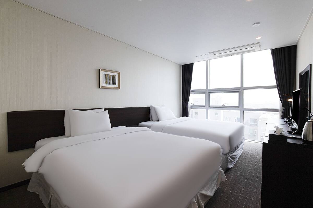 Quarto de hotel com duas camas, uma janela grande, um quadro na parede e uma mesa com abajur.