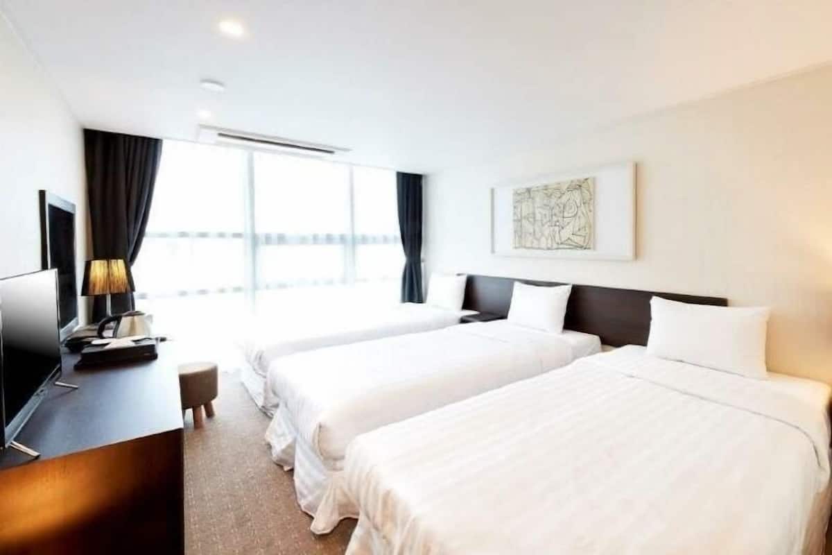 Quarto de hotel com duas camas, uma escrivaninha com computador, televisão e janela com cortinas.