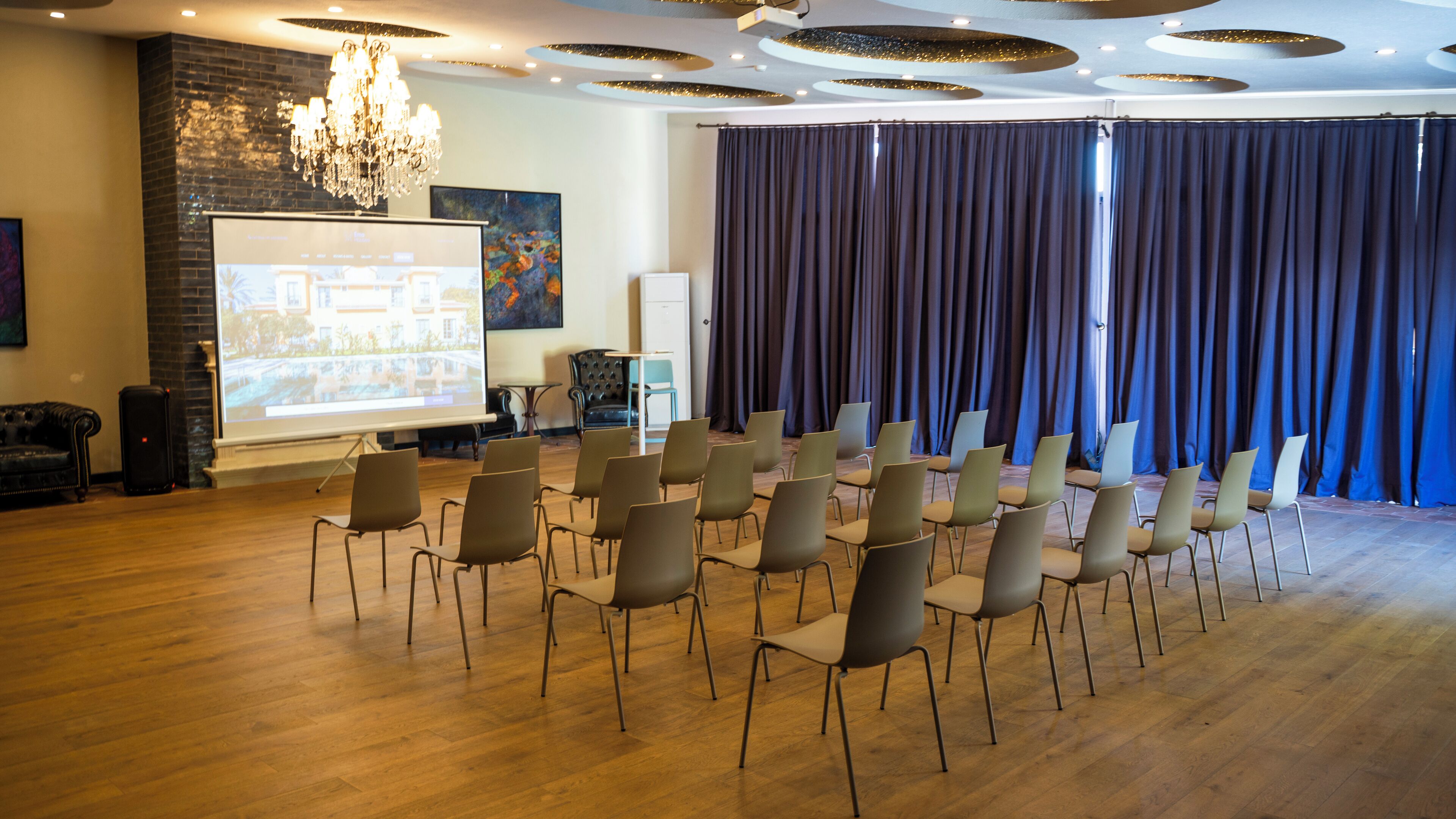 Uma sala de conferências com cadeiras dispostas em fileiras, uma tela de projetor, um candelabro e uma janela grande com cortinas.
