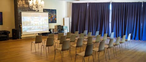 Uma sala de conferências com cadeiras dispostas em fileiras, uma tela de projetor, um candelabro e uma janela grande com cortinas.