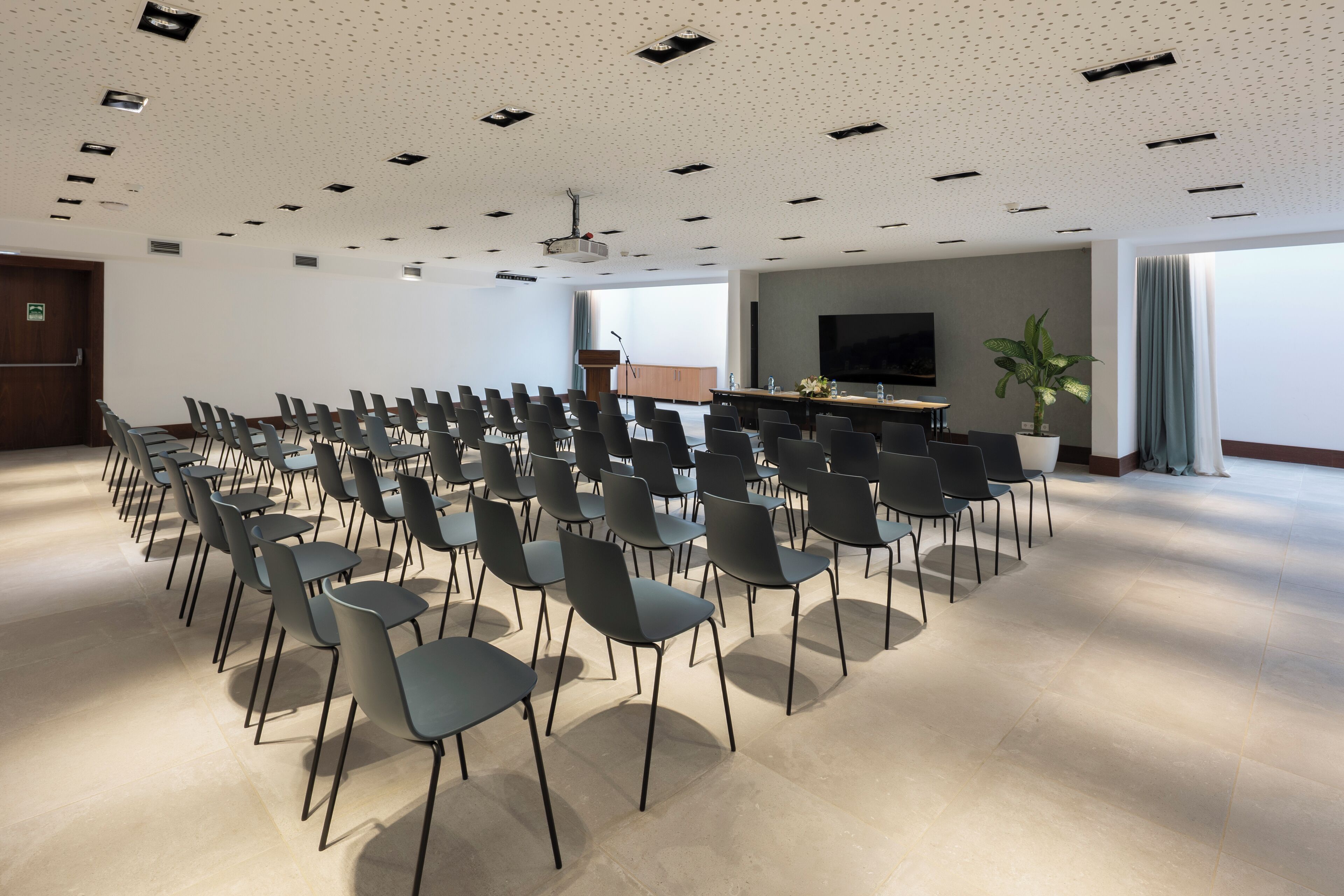 Sala de conferência com fileiras de cadeiras, um pódio, uma tela grande e um vaso de planta.