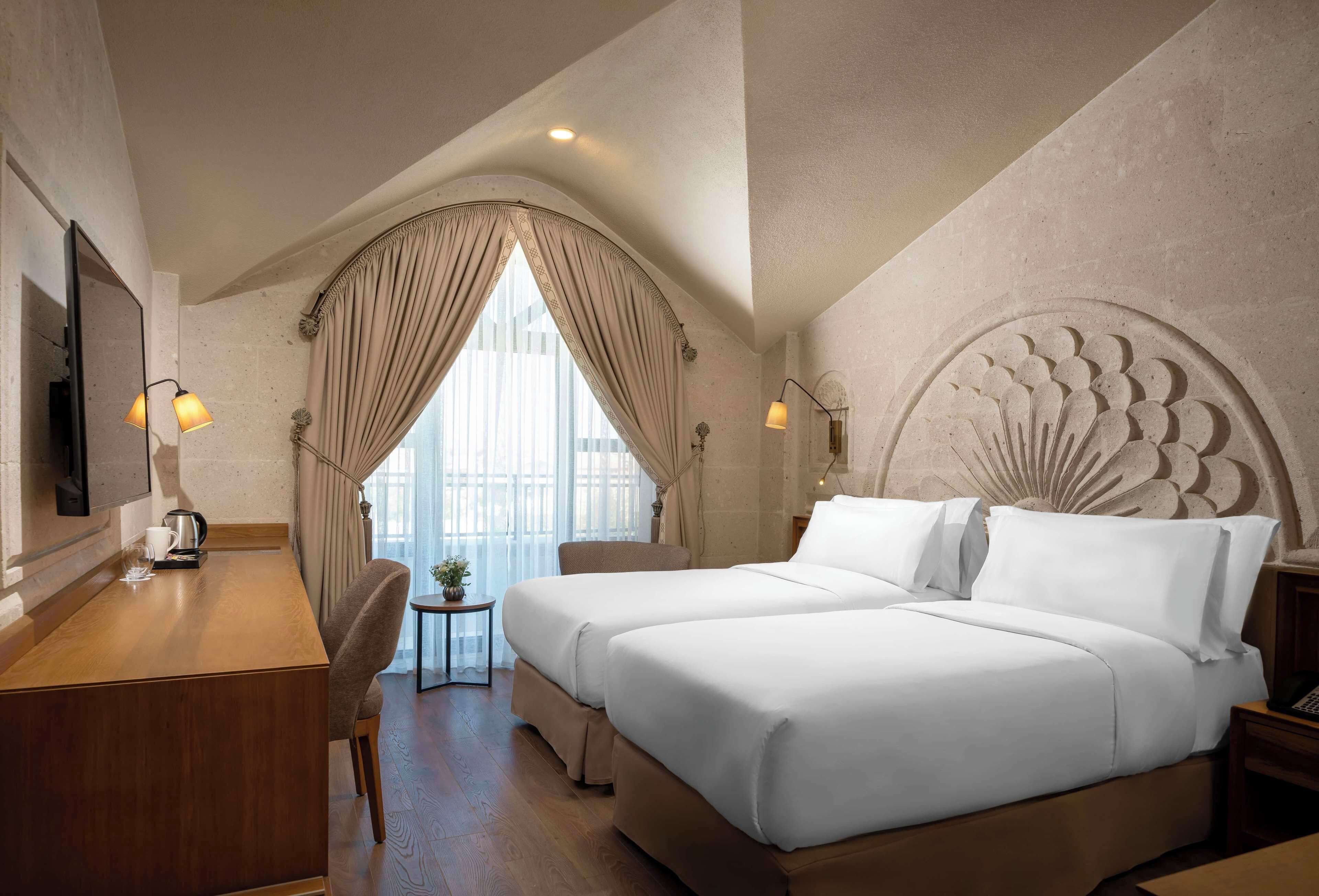 Quarto de hotel com duas camas, uma escrivaninha, uma cadeira e uma pequena mesa com uma planta.