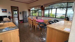 Uma área de refeições com mesa e cadeiras de madeira, cozinha com geladeira e grandes janelas com vista para vegetação e um corpo d'água.