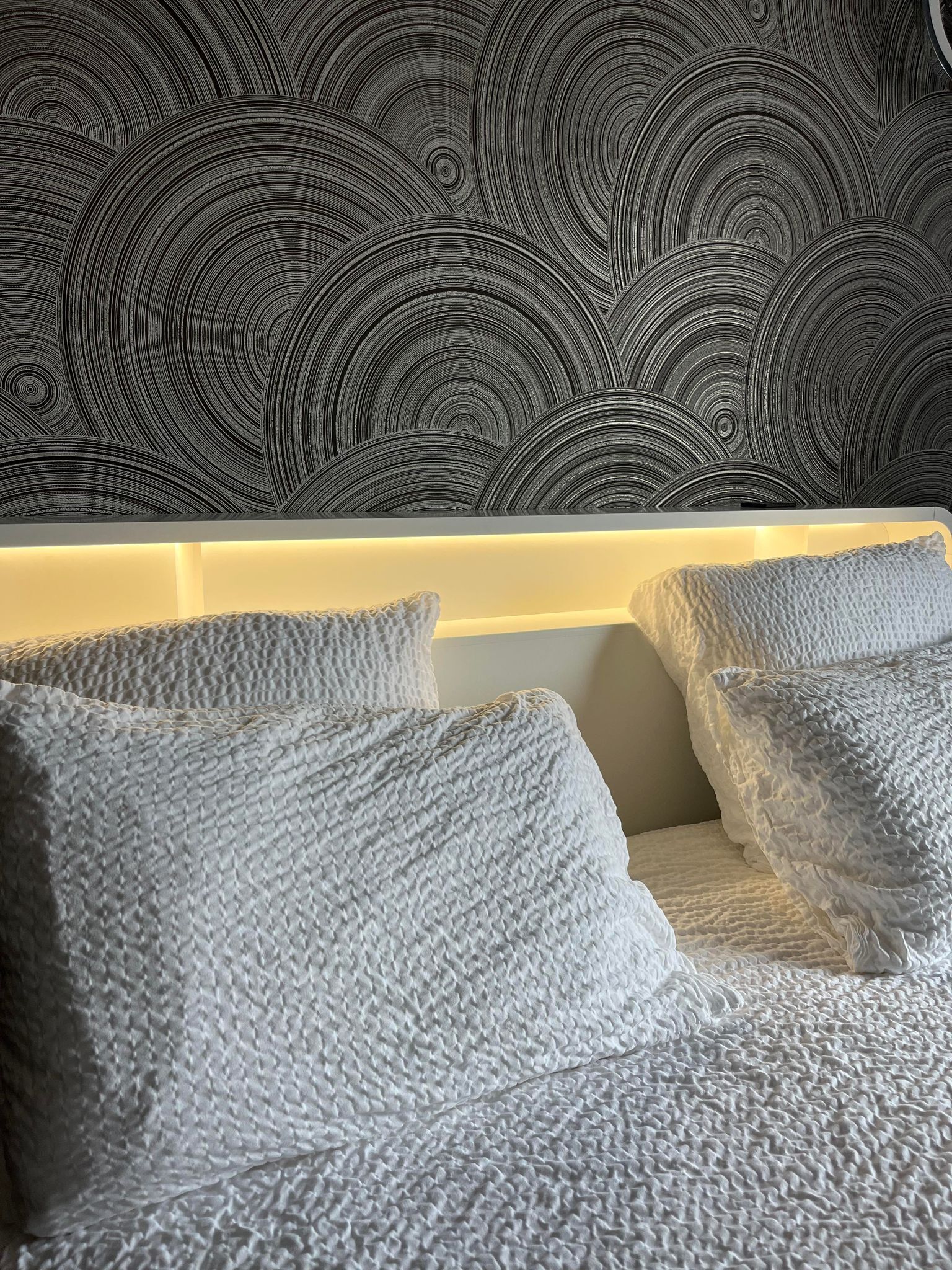 Cama com almofadas brancas texturizadas e cabeceira com padrão circular.