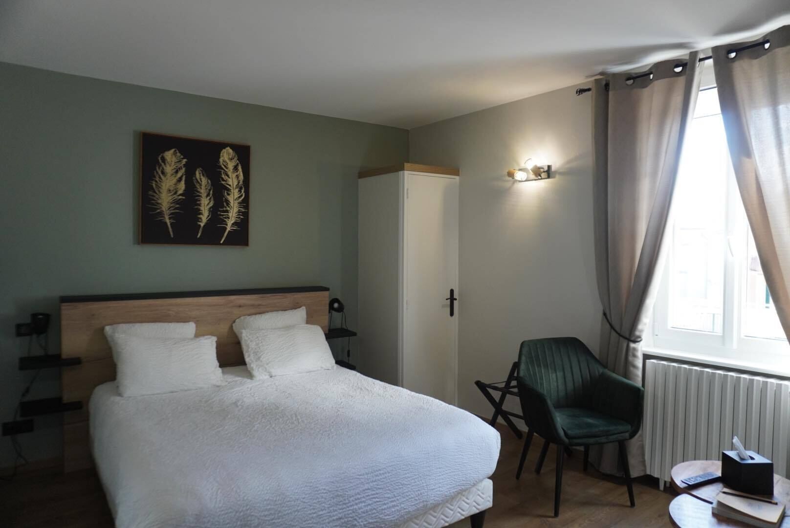 Quarto de hotel com cama, poltrona, escrivaninha, radiador e janela com cortinas.