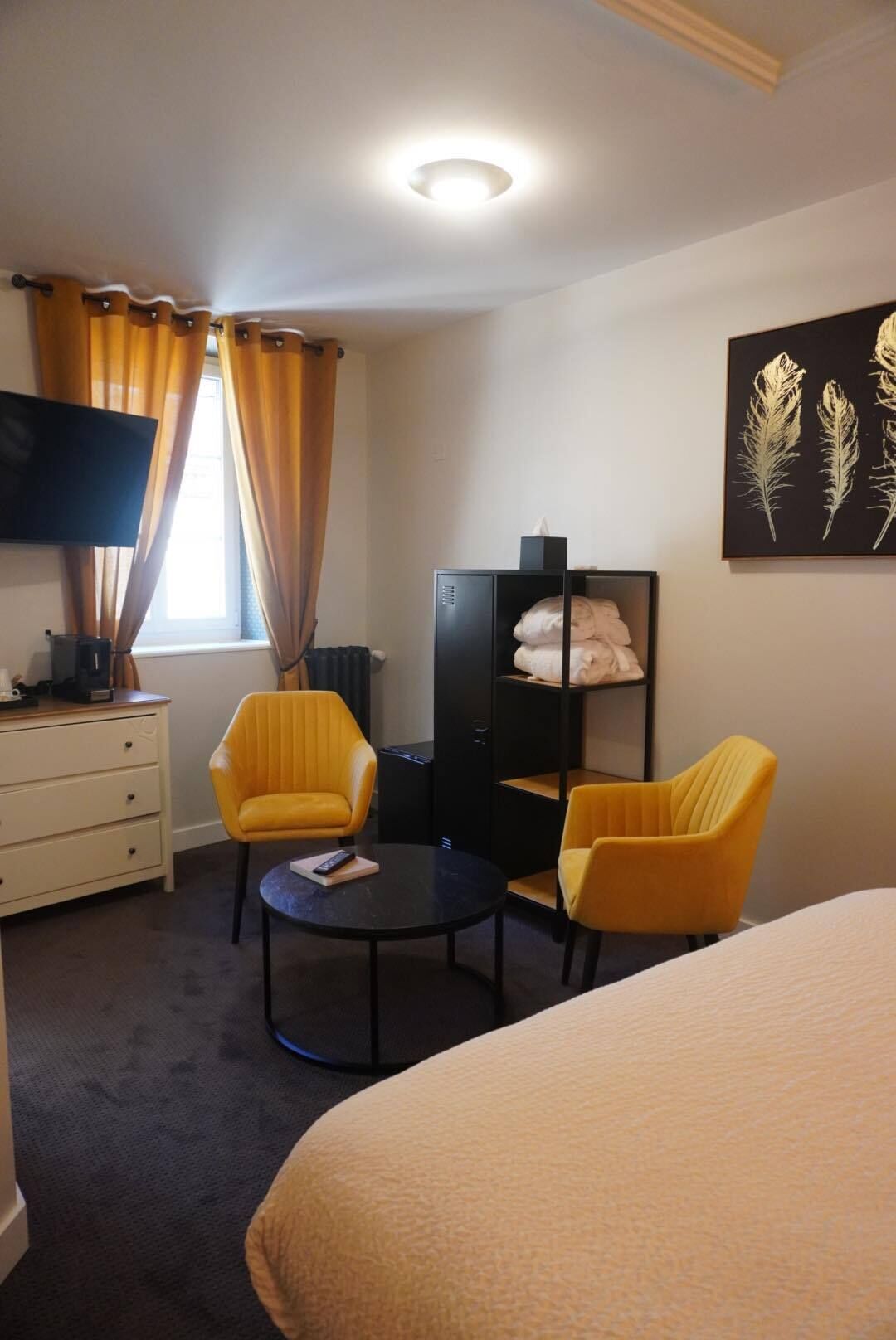 Quarto de hotel com uma cama, duas poltronas amarelas, uma mesa redonda pequena, uma televisão, um guarda-roupa e um quadro na parede.