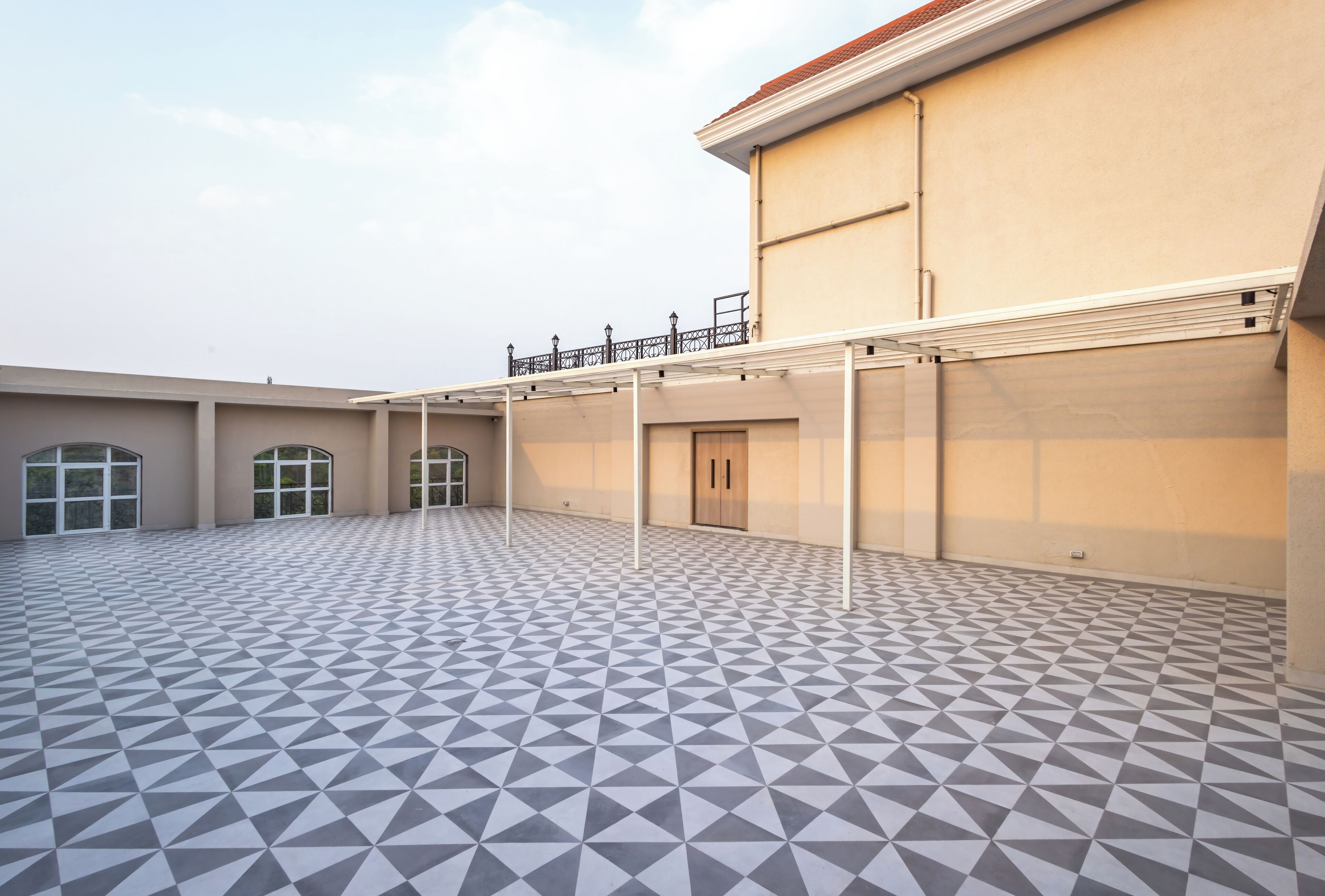 Um pátio amplo com piso de azulejos geométricos, grandes janelas e um edifício com fachada bege e telhado de telhas vermelhas.