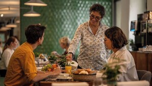 Uma mesa posta para três pessoas, com cesta de pão e uma garrafa de suco de laranja.