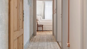 Um corredor com piso de madeira, uma cama com roupa de cama branca e uma cadeira.