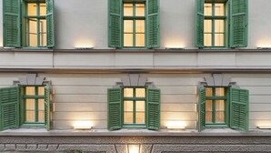 Fachada de edifício com várias janelas, persianas verdes, luminárias espaçadas uniformemente e base de pedra.