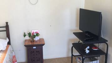 Quarto com cama, uma pequena mesa de cabeceira de madeira, televisão em móvel e prateleira na parede com uma fotografia.