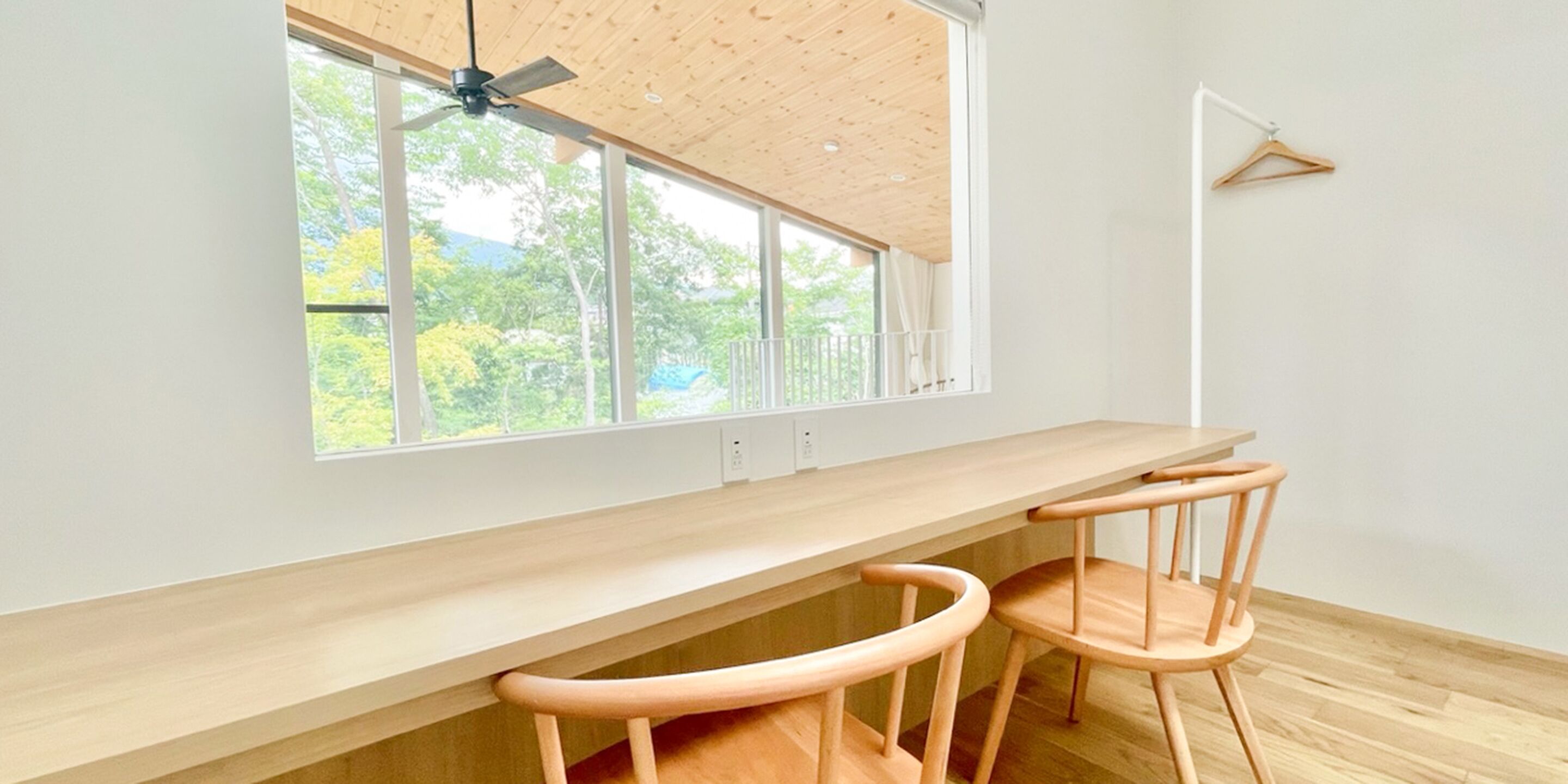 Um quarto com uma longa mesa de madeira, duas cadeiras de madeira, um ventilador de teto e uma grande janela com vista para árvores e uma construção azul lá fora.