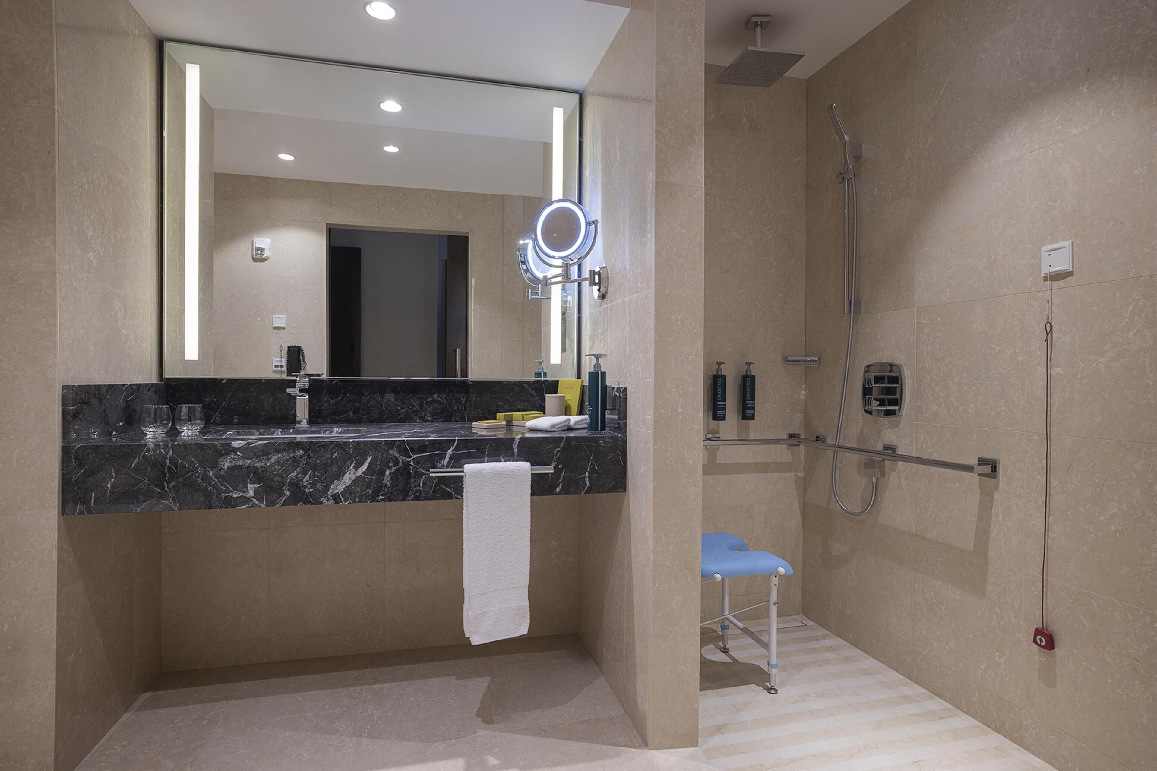 Um banheiro moderno com bancada de mármore, um espelho grande, chuveiro manual e um banquinho azul.