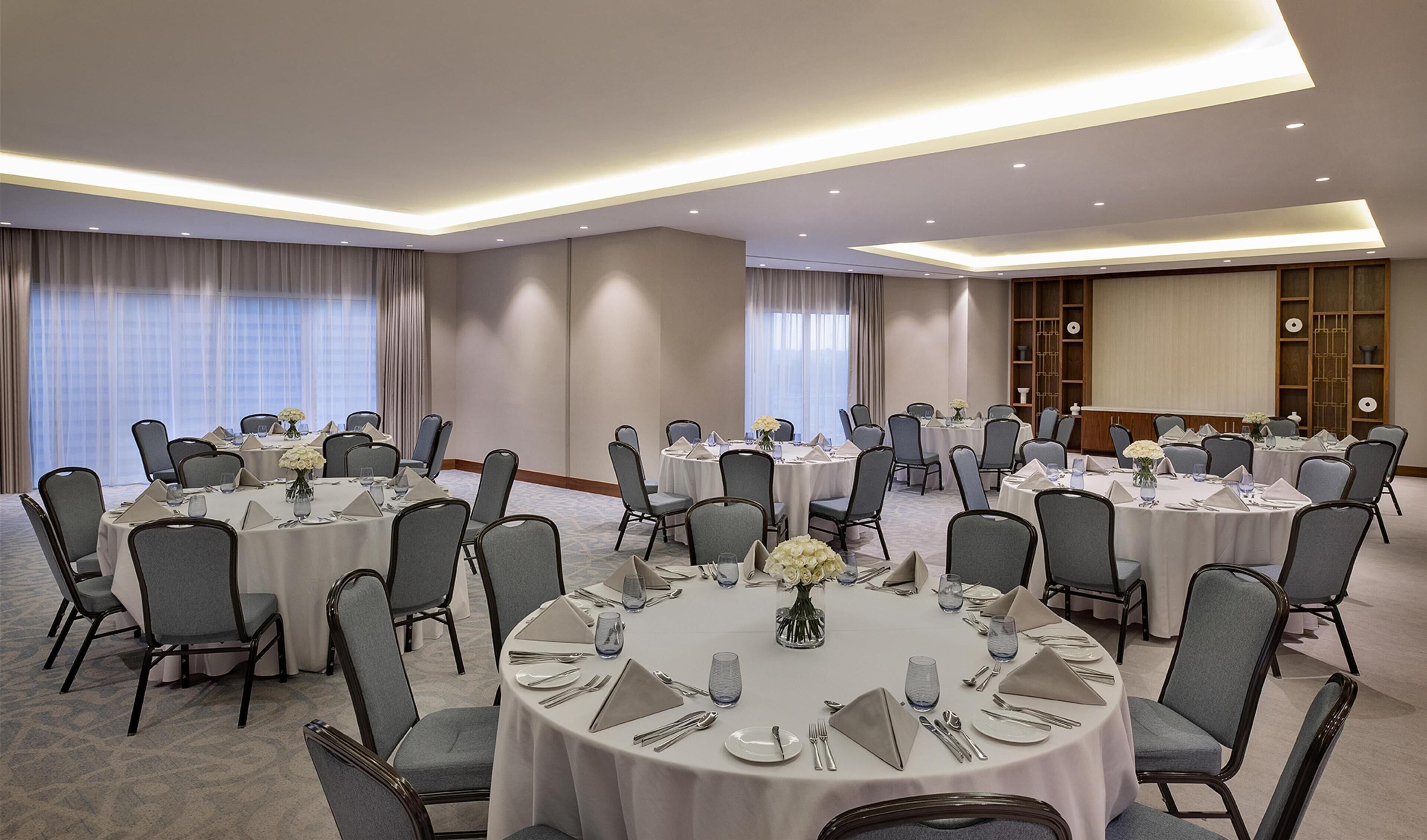 Sala de conferências com mesas redondas, cadeiras cinzas e toalhas de mesa brancas.