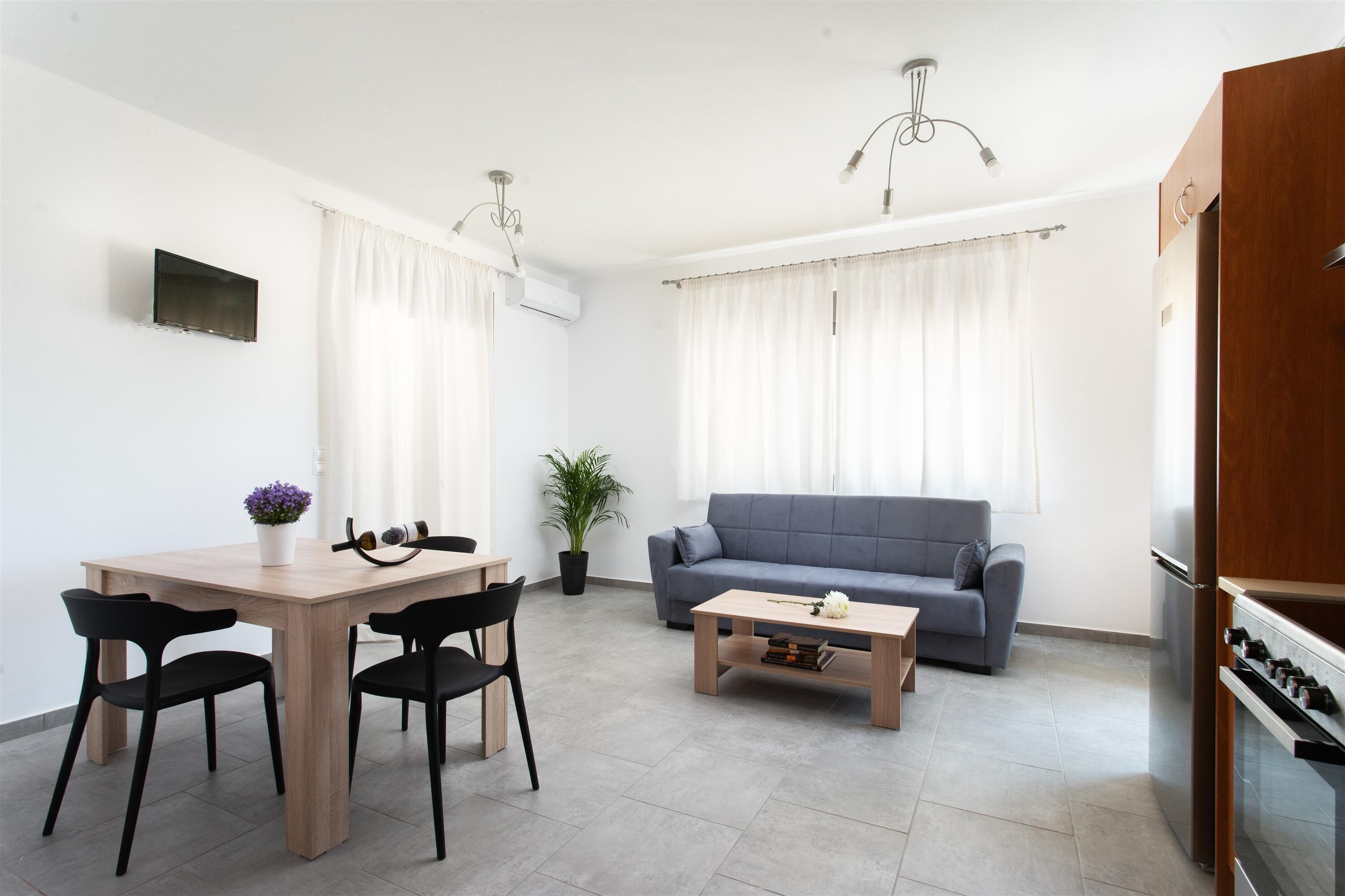 Uma sala moderna com um sofá cinzento, uma mesa de jantar de madeira com quatro cadeiras, uma mesa de centro, uma TV fixada na parede e uma área de cozinha com fogão.