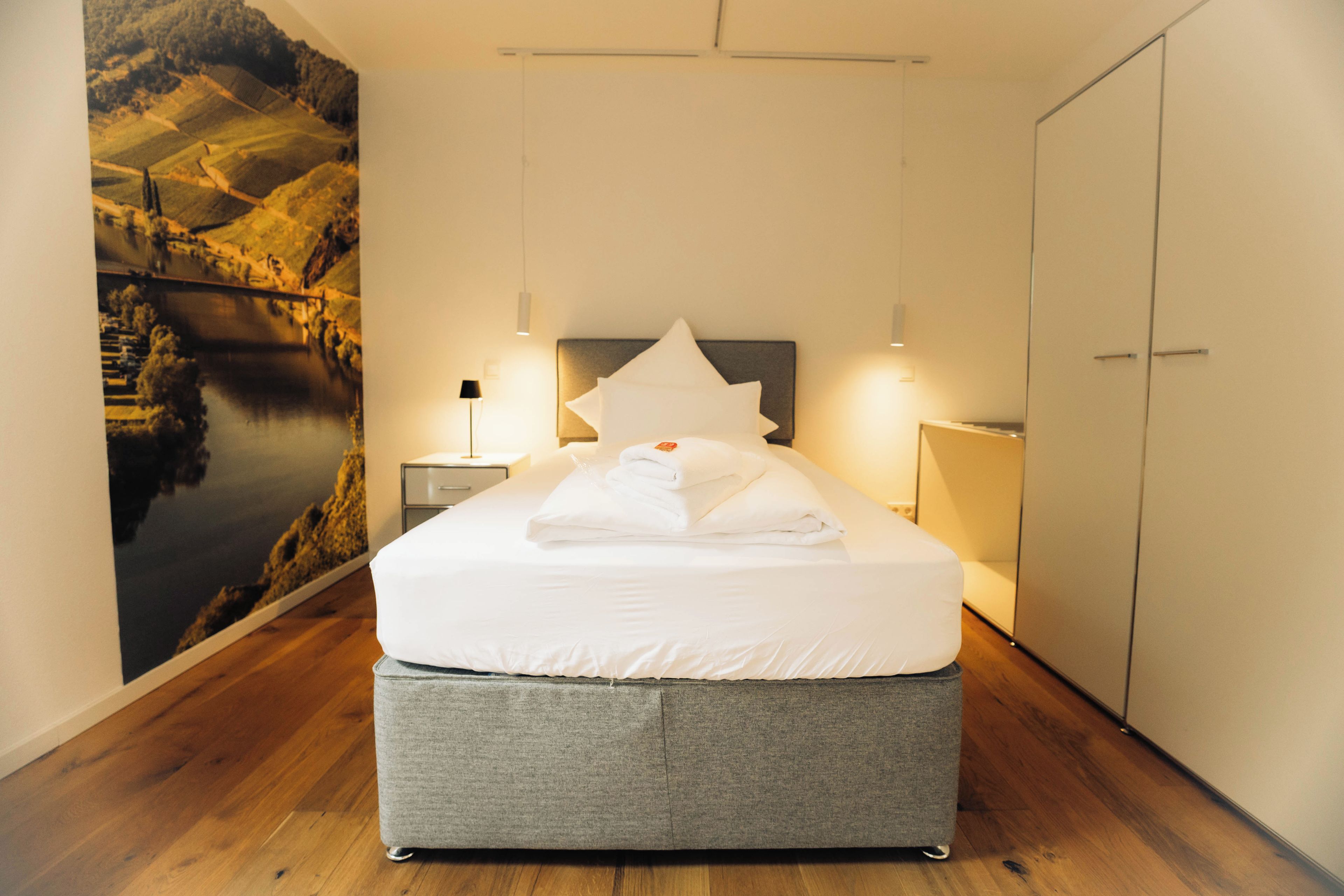 Cama bem arrumada com lençóis brancos em quarto moderno de hotel, com um mural grande na parede, mesa de cabeceira com abajur e guarda-roupa.