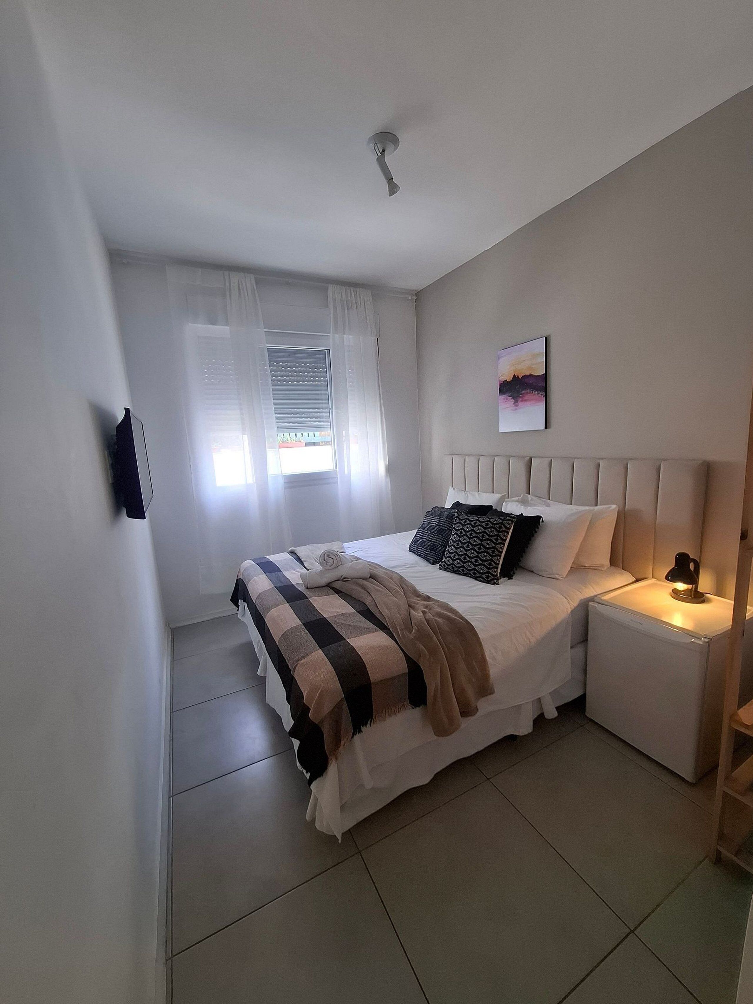 Quarto com cama, mesa de cabeceira com abajur, janela com cortinas e uma obra de arte na parede.