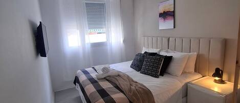 Quarto com cama, mesa de cabeceira com abajur, janela com cortinas e uma obra de arte na parede.