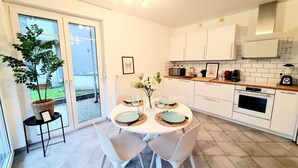 Uma cozinha moderna com armários brancos, uma área de jantar com mesa e cadeiras redondas, e uma janela grande com vista para o exterior.