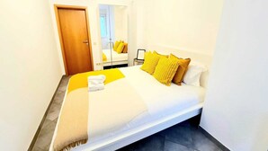 Quarto de hotel com uma cama, um espelho, uma porta e uma pequena mesa com um porta-retrato.