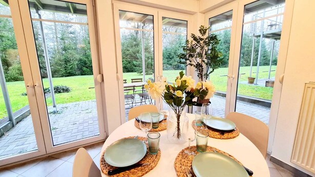 Uma sala de jantar com uma mesa redonda posta para quatro pessoas, cercada por grandes janelas com vista para um jardim.