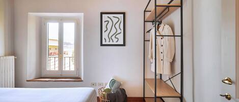 Quarto com cama, janela, radiador, prateleira com roupas e quadro emoldurado.