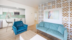 Uma sala de estar moderna com um sofá e poltrona azuis, uma mesa de centro de madeira e uma cozinha ao fundo.