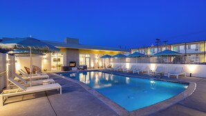 Piscina com espreguiçadeiras e guarda-sóis, rodeada por um edifício com cerca branca e telhado plano.