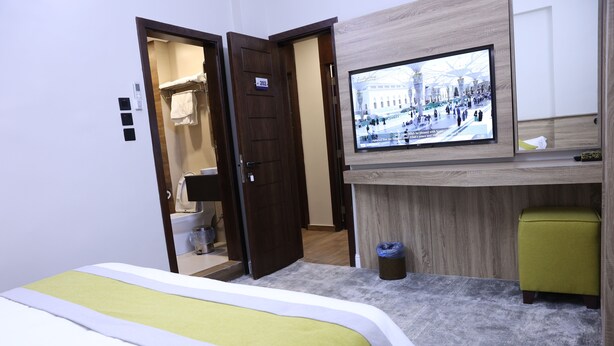 Quarto de hotel com cama, televisão fixada na parede, secretária de madeira, um banquinho verde e banheiro visível por uma porta aberta.