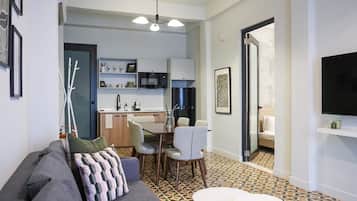 Uma sala de estar moderna com um sofá cinzento, uma mesa de jantar com cadeiras, uma cozinha com micro-ondas e pia, e uma TV na parede.
