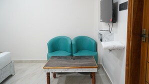 Um quarto pequeno com um sofá branco, um sofá verde-água, uma mesa com tampo de mármore, uma TV fixada na parede e uma porta de madeira.