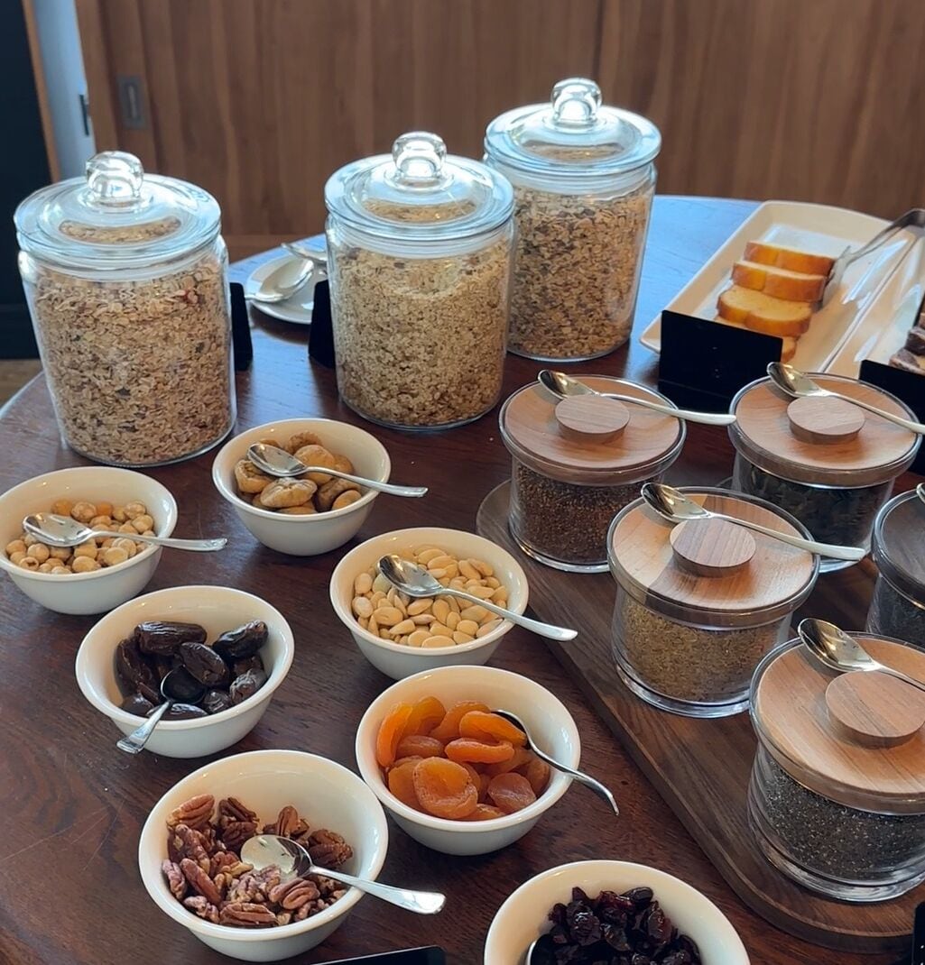 Uma mesa de buffet com diversos alimentos e recipientes, incluindo tigelas com nozes, frutas secas e cereais.