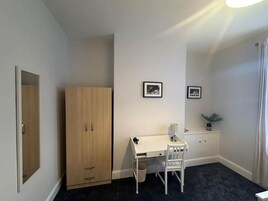 Quarto com um guarda-roupa de madeira, uma escrivaninha branca com cadeira e duas imagens emolduradas na parede.