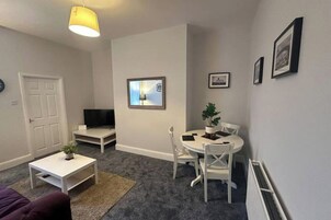 Um espaço de estar compacto com porta branca, televisão de tela plana, mesa pequena branca, mesa de jantar com cadeiras e um vaso de planta.