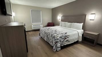 Quarto de hotel com cama, mesas de cabeceira, televisão, janela com persianas e uma cadeira vermelha.