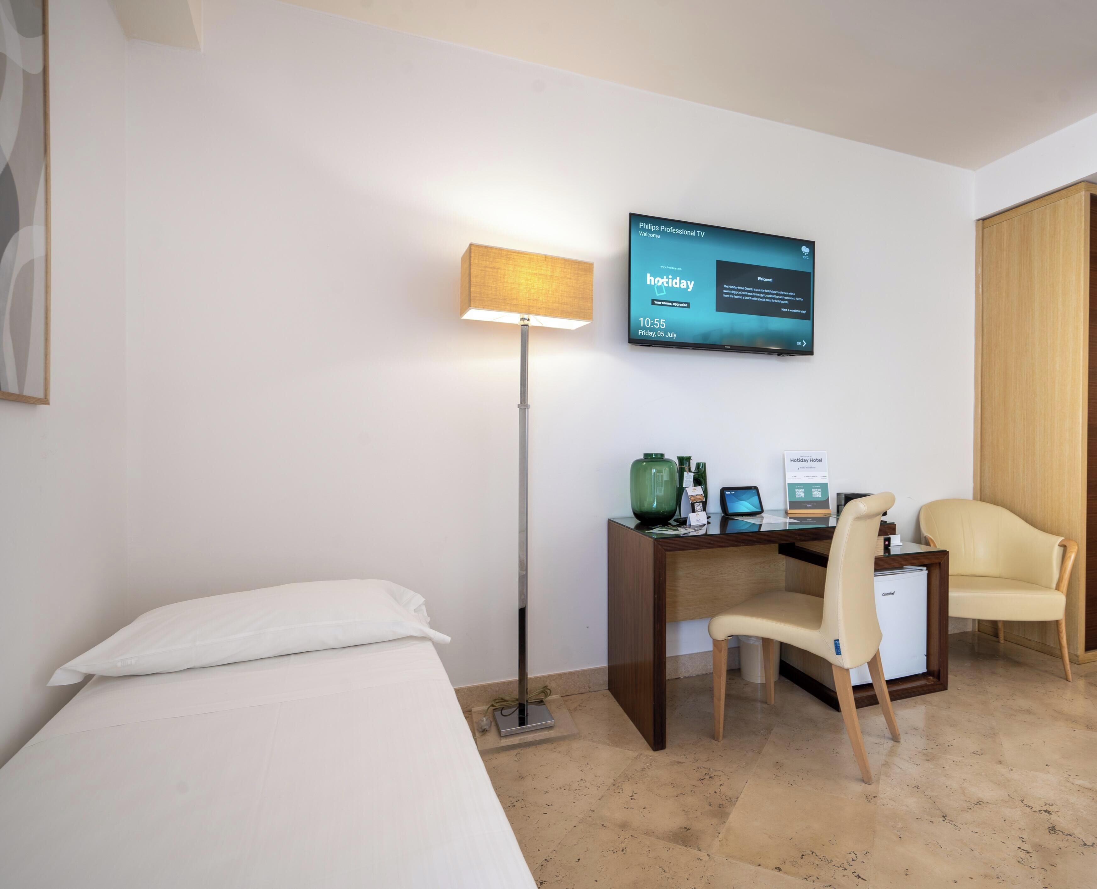 Quarto de hotel com cama, escrivaninha com computador, cadeira, luminária e televisão exibindo uma mensagem de boas-vindas.