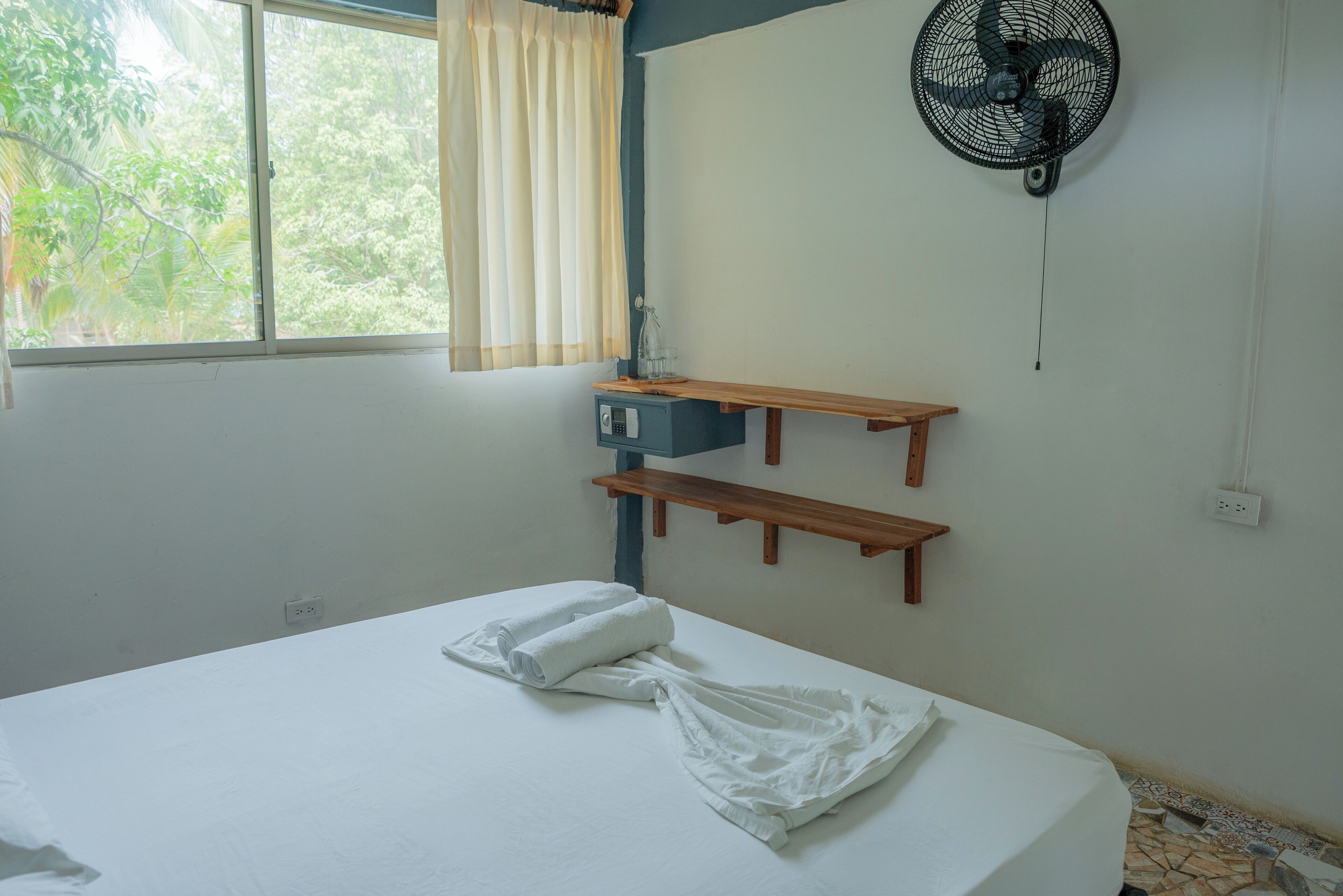 Quarto de hotel com cama, janela com cortinas, ventoinha fixada na parede e prateleiras de madeira.