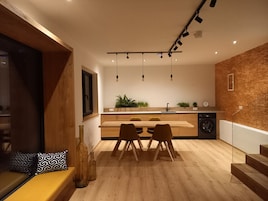 Uma cozinha moderna com armários de madeira, forno embutido e pia. Há um espaço de refeições com mesa e cadeiras de madeira, além de um banco com almofadas.
