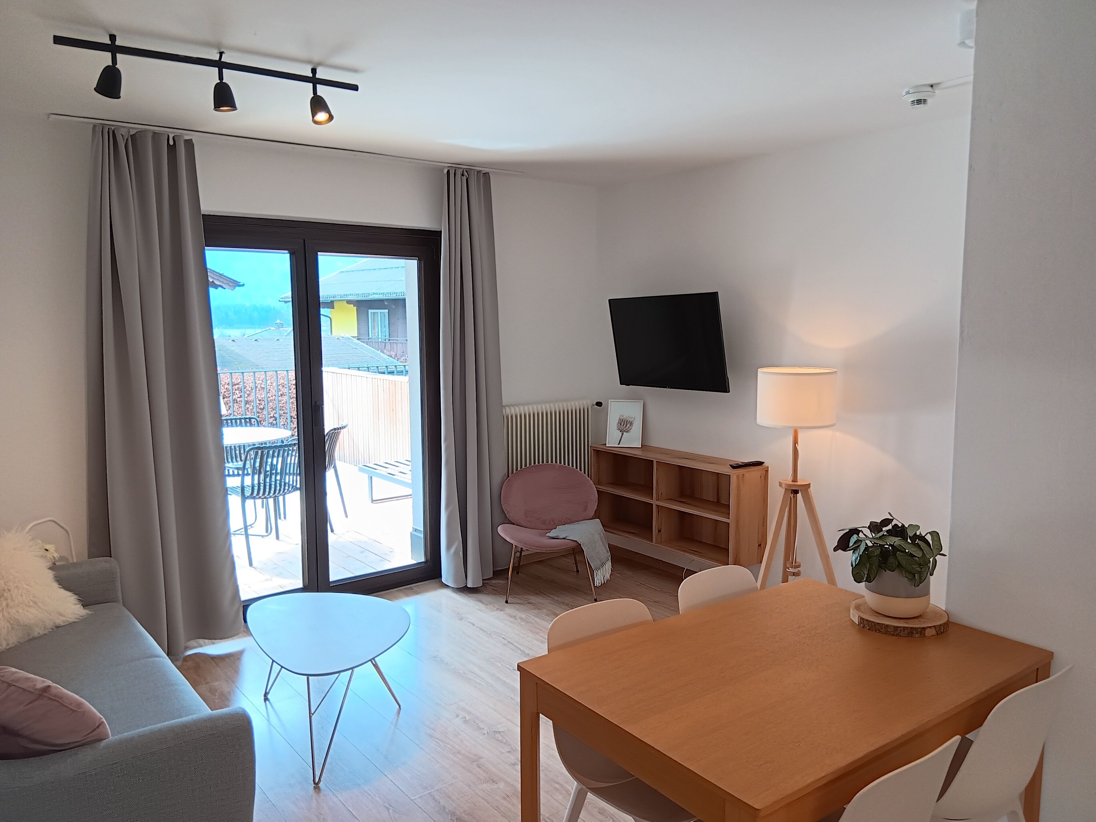 Uma sala de estar moderna com um sofá, uma mesa pequena, uma mesa de jantar de madeira, uma TV fixada na parede e um vaso com planta.