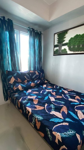 Uma cama de solteiro com colcha estampada em folhas num quarto com janela, cortinas e um quadro pendurado na parede.
