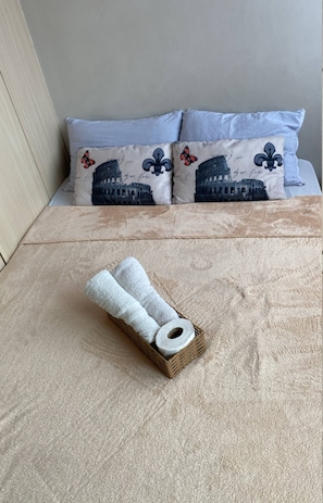 Cama com duas almofadas de padrão com um edifício e borboleta, tapete bege texturizado e cesta com toalhas dobradas.
