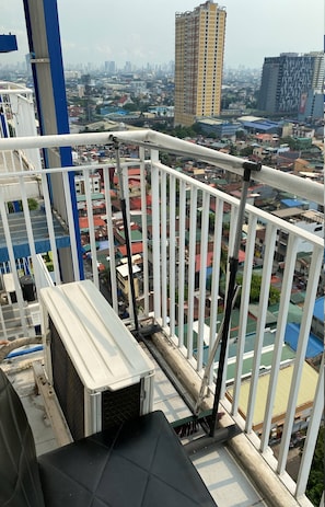 Uma varanda com grade de metal, um aparelho de ar condicionado branco e vista para um horizonte urbano com edifícios.