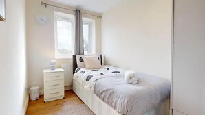Um quarto pequeno e bem arrumado, com uma cama de solteiro, uma mesinha de cabeceira e uma janela.