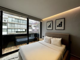 Quarto moderno com uma cama grande, um pequeno espaço para sentar e vista para um edifício com varandas.