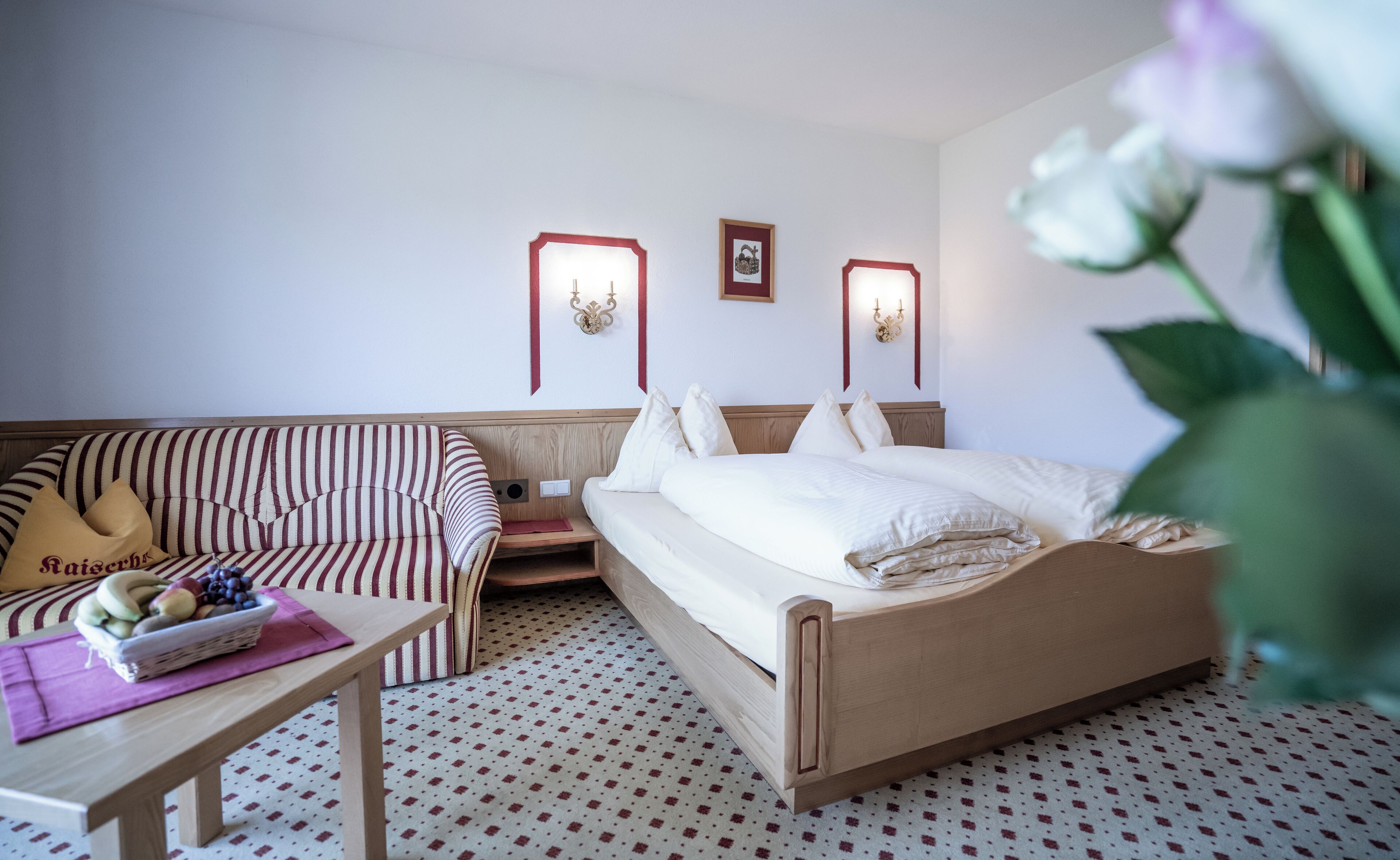 Quarto de hotel com sofá listrado, cama com roupa de cama branca, mesa pequena com cesta de frutas e duas luminárias fixadas na parede.